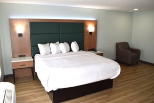 Quarto de hotel com uma cama grande, duas mesas de cabeceira, uma cadeira e cabeceira com iluminação embutida.