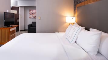 Quarto de hotel com uma cama grande, uma televisão em um suporte de madeira, uma escrivaninha com gavetas, uma cadeira e uma luminária fixada na parede.
