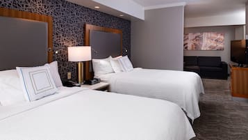 Quarto de hotel com duas camas, uma escrivaninha com abajur, televisão e sofá.