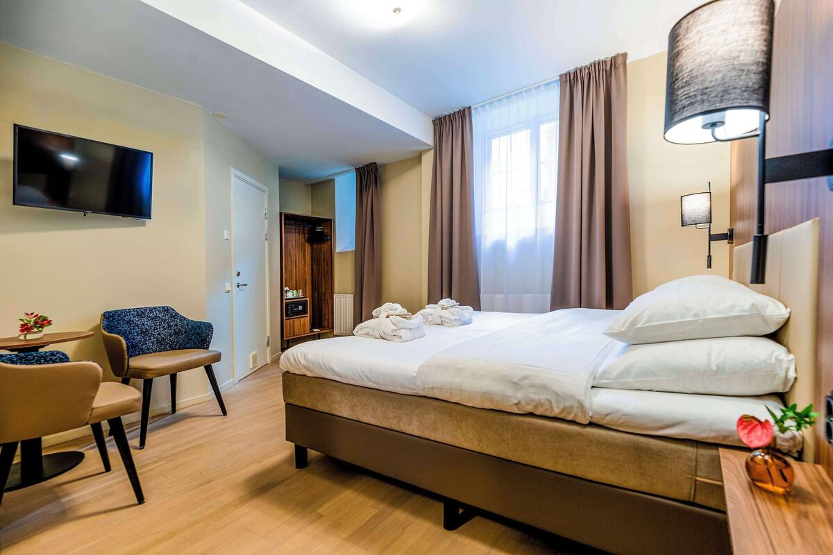 Quarto de hotel com cama, escrivaninha, cadeira, televisão e luminária.