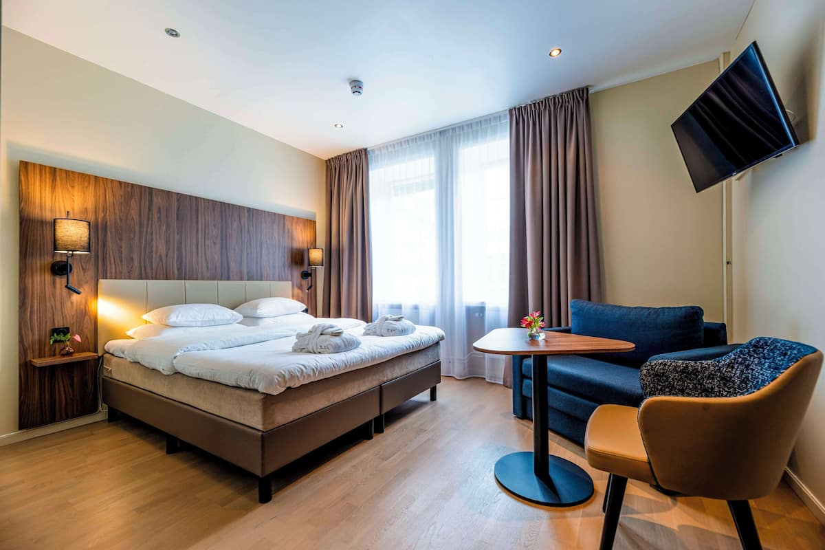 Quarto de hotel moderno com uma cama grande, uma mesinha, um sofá e uma televisão presa à parede.