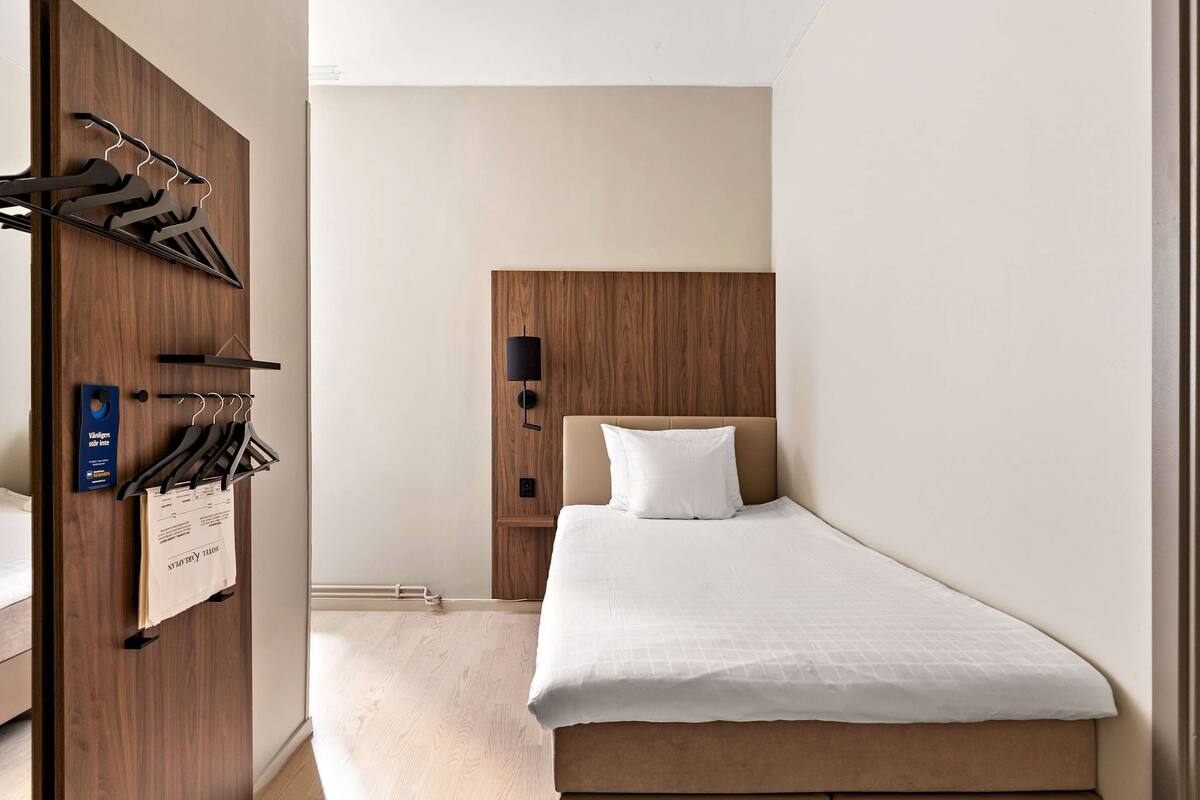 Quarto de hotel com cabeceira de madeira, cama com roupa de cama branca, cabideiro fixado na parede e mesa de cabeceira.
