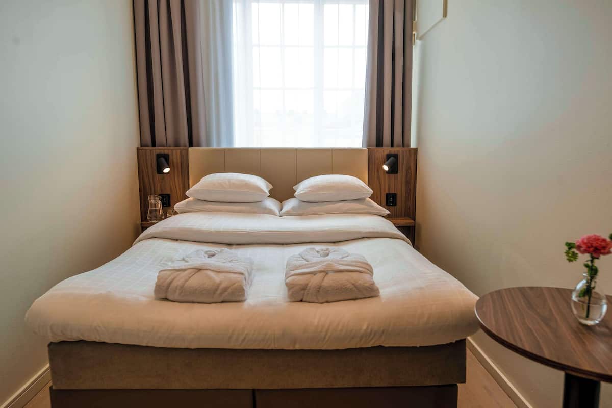 Cama bem arrumada com lençóis brancos e duas toalhas, mesa de cabeceira com um vaso de flores e uma janela com cortinas.