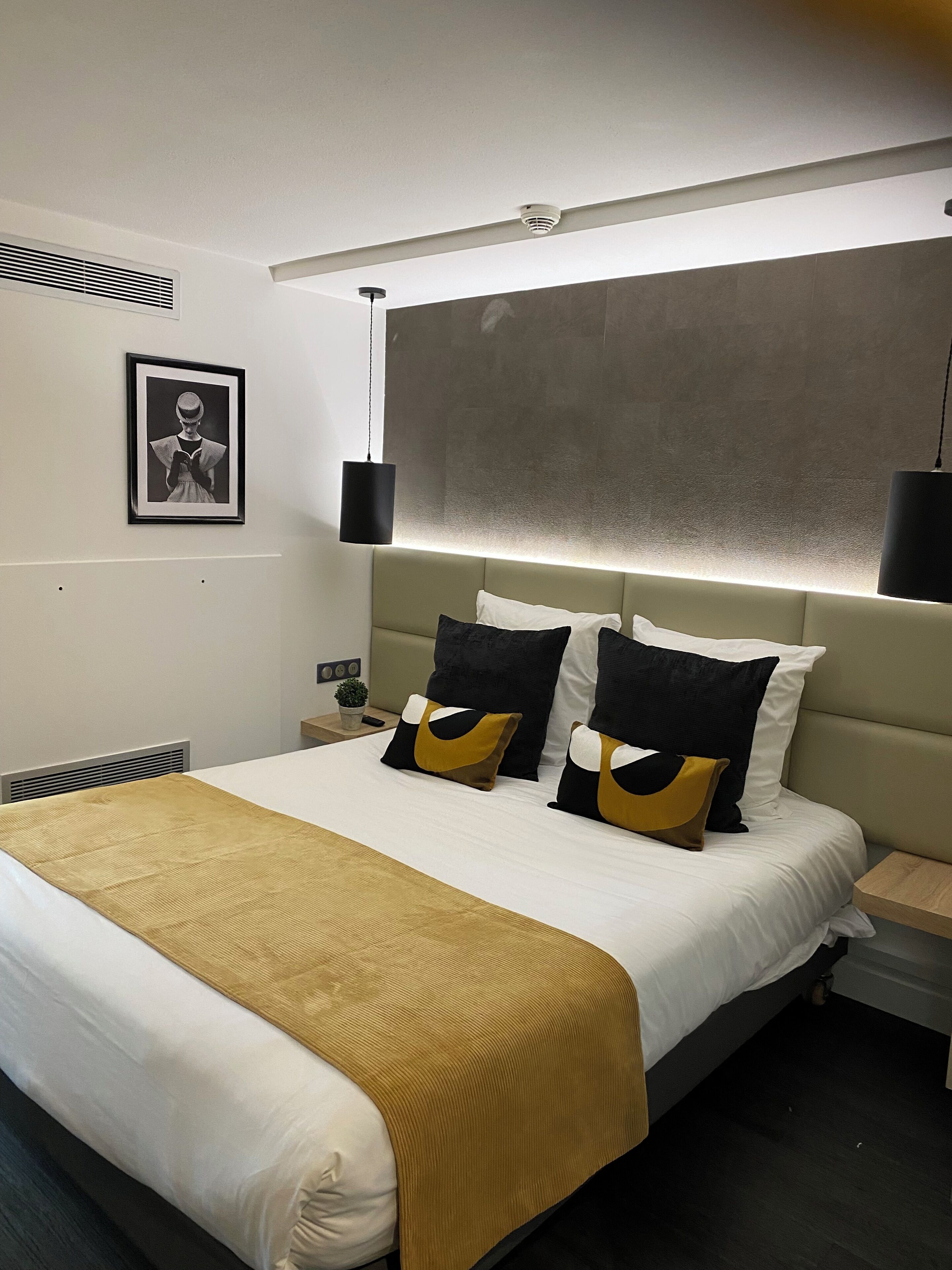 Quarto de hotel moderno com uma cama grande, cabeceira com iluminação integrada, quadro na parede e mesa de cabeceira com abajur.