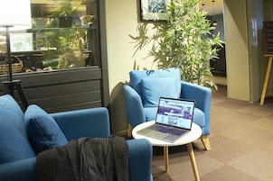 Uma área de espera moderna com poltronas azuis, um laptop sobre uma mesa pequena e uma grande planta em vaso.