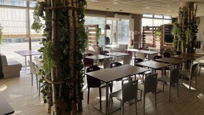 Espaço interior moderno com mesas, cadeiras, janelas amplas e instalações de plantas decorativas.