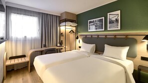 Quarto de hotel com duas camas, uma mesa, uma cadeira, uma mesinha, um abajur e duas imagens emolduradas na parede.
