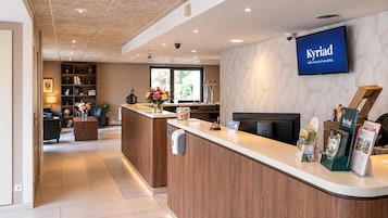 Uma área de recepção de hotel com balcão de madeira, televisão fixada exibindo o nome do hotel e vista do saguão com assentos e lareira.