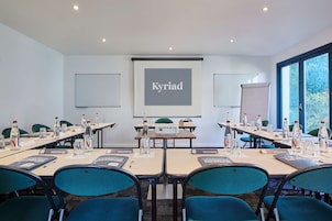 Sala de conferência com tela de projetor exibindo 'Kyriad', mesas com garrafas de água, placas de identificação e cadeiras.