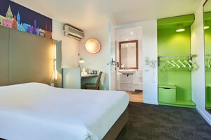 Quarto de hotel com cama, escrivaninha, cadeira, espelho, pia e parede verde com porta-toalhas.
