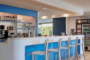 Uma área de bar com esquema de cores azul e branco, banquetas, prateleiras com garrafas e um mini frigorífico.