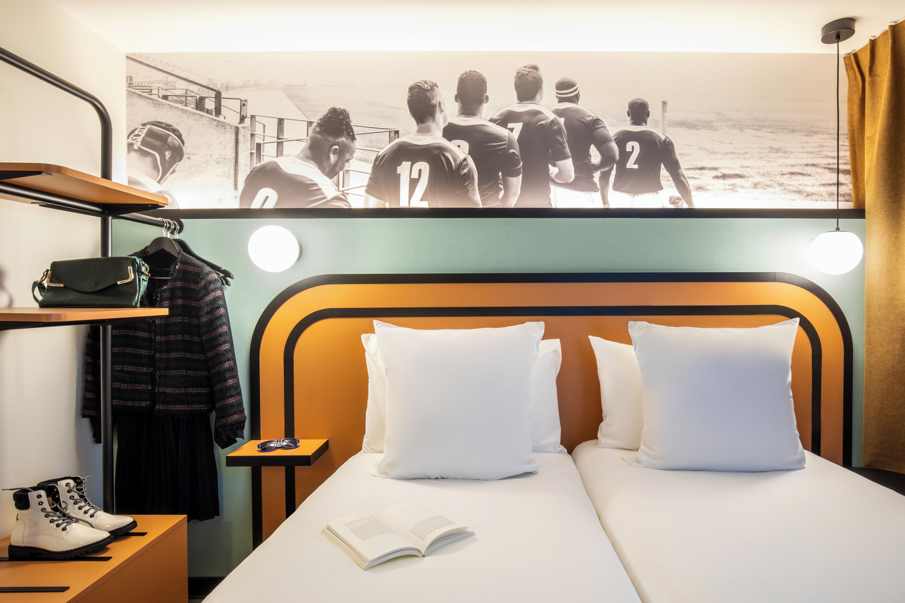 Quarto de hotel com uma cama, almofadas, um livro, um cabideiro com um casaco e uma parede com um pôster.