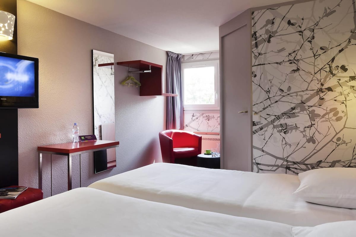 Quarto de hotel com cama, escrivaninha com cadeira, televisão, janela com cortinas e parede com padrão floral.