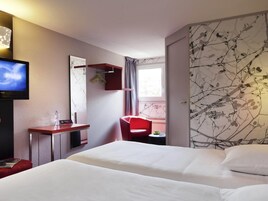 Quarto de hotel com cama, escrivaninha com cadeira, televisão, janela com cortinas e parede com padrão floral.