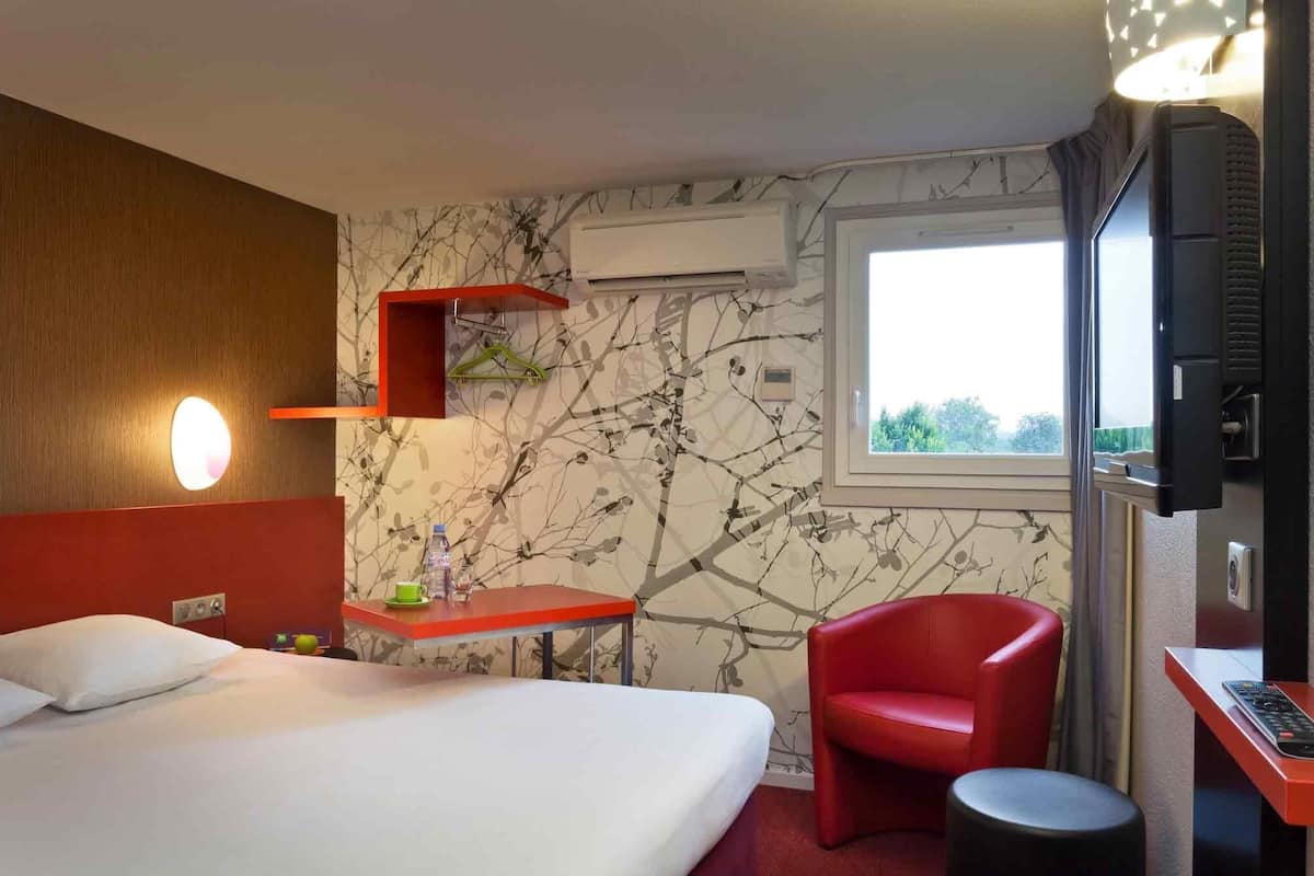 Quarto de hotel com cama, uma cadeira vermelha, mesa pequena, TV fixada na parede e ar condicionado.