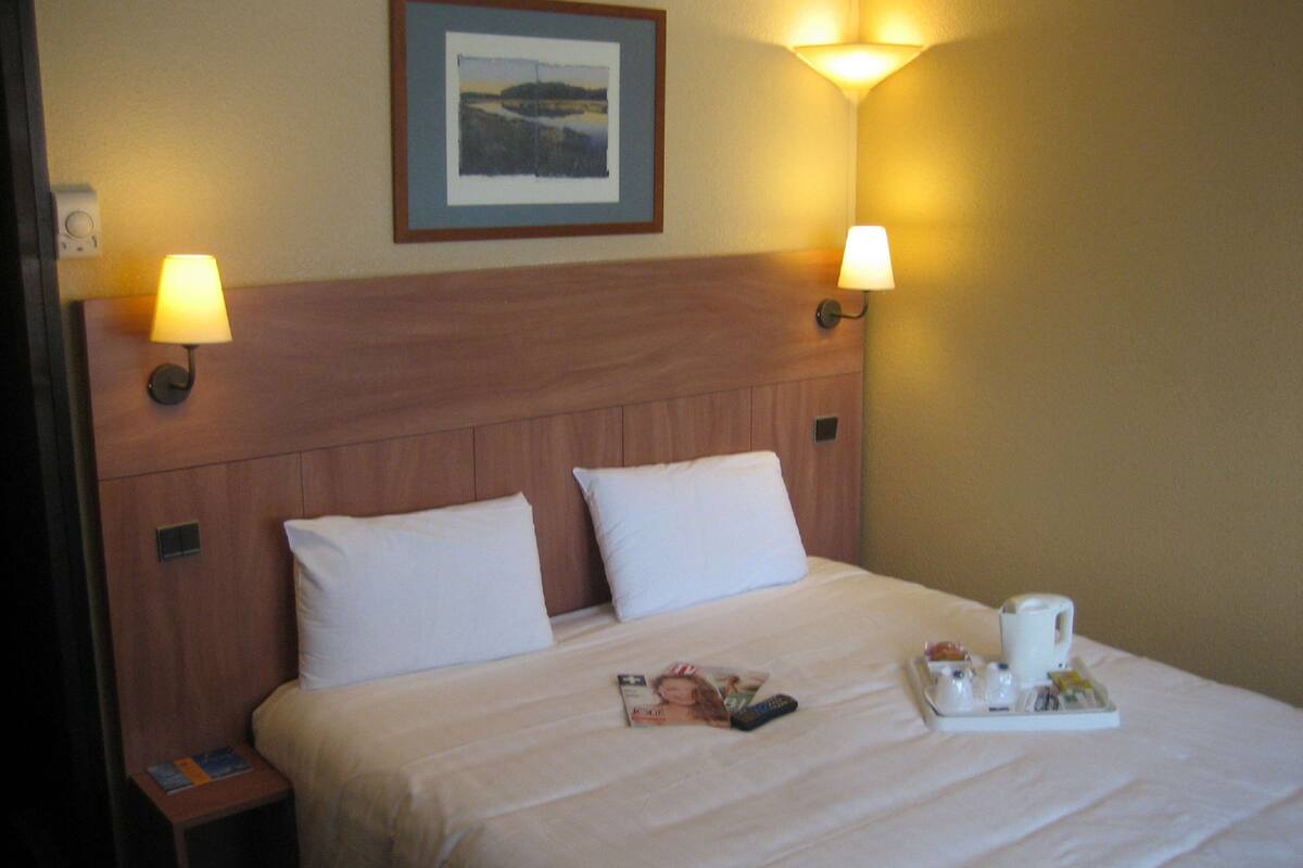 Quarto de hotel com uma cama, duas luminárias de cabeceira, um quadro na parede e um tabuleiro com uma cafeteira e outros objetos.