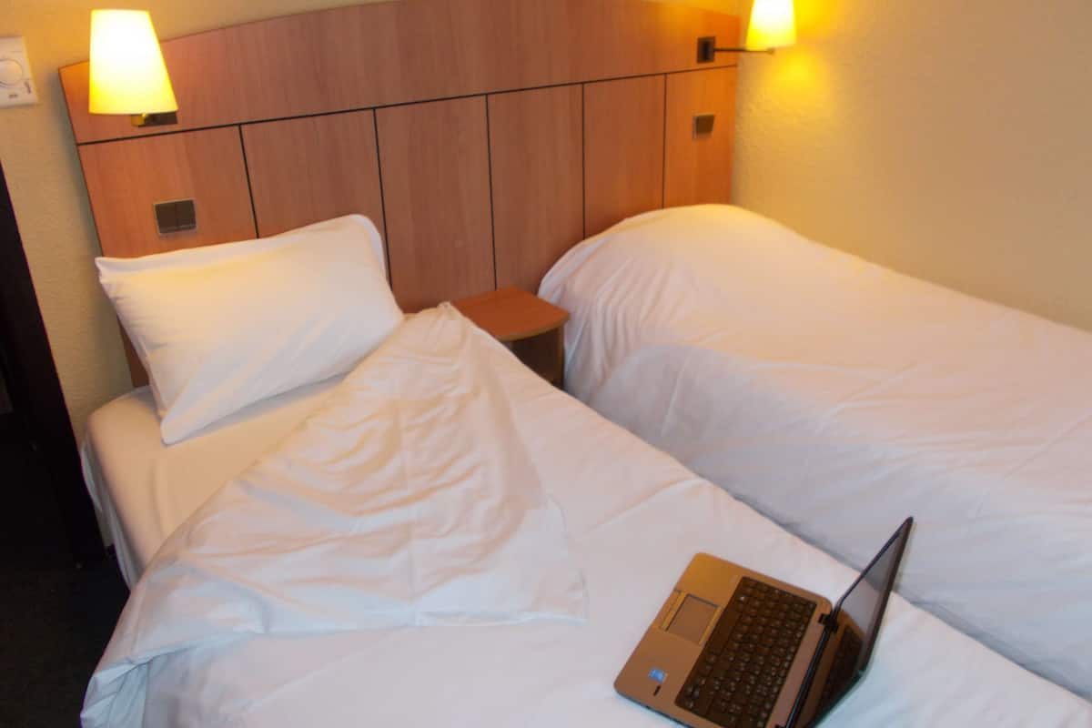 Quarto de hotel com duas camas, uma cabeceira de madeira, luminárias de cabeceira e um laptop sobre uma das camas.