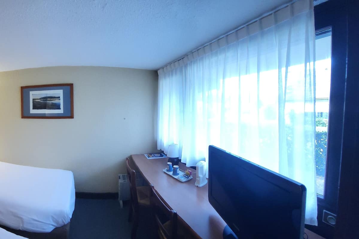 Quarto de hotel com duas camas de solteiro, uma escrivaninha com computador, uma janela com cortinas e um quadro na parede.