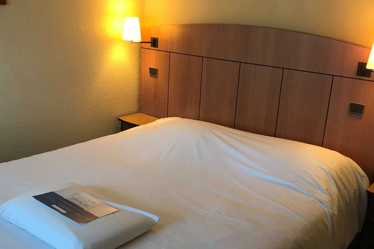 Cama bem arrumada com travesseiro e um cartão, cabeceira de madeira e duas luminárias fixadas na parede.