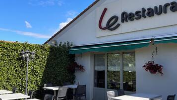 Uma área externa com mesas e cadeiras, um edifício com uma placa indicando 'Le Restaurante' e um arbusto ao fundo.