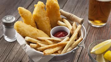 Um balde de peixe com batatas fritas, um pequeno copo de ketchup, um copo de cerveja e um saleiro sobre uma mesa de madeira.