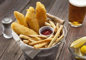 Um balde de peixe com batatas fritas, um pequeno copo de ketchup, um copo de cerveja e um saleiro sobre uma mesa de madeira.