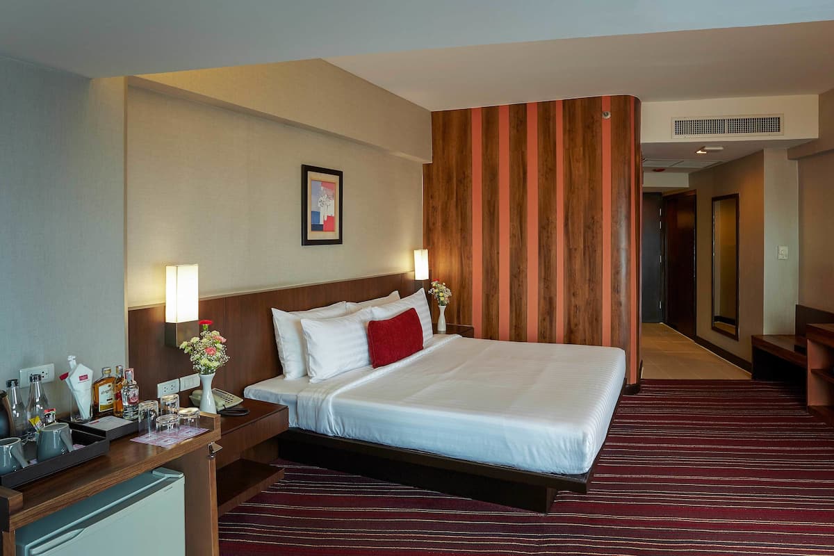 Quarto de hotel com uma cama grande, uma parede de destaque em madeira, uma escrivaninha com abajur e uma mesinha com um vaso de flores.