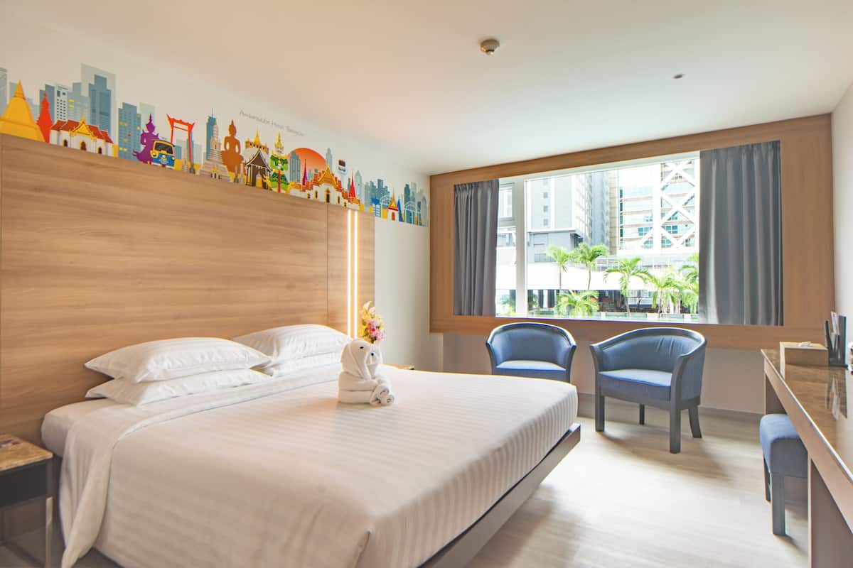Quarto de hotel com cama grande, duas cadeiras, escrivaninha, mural com vista urbana e janela com cortinas.