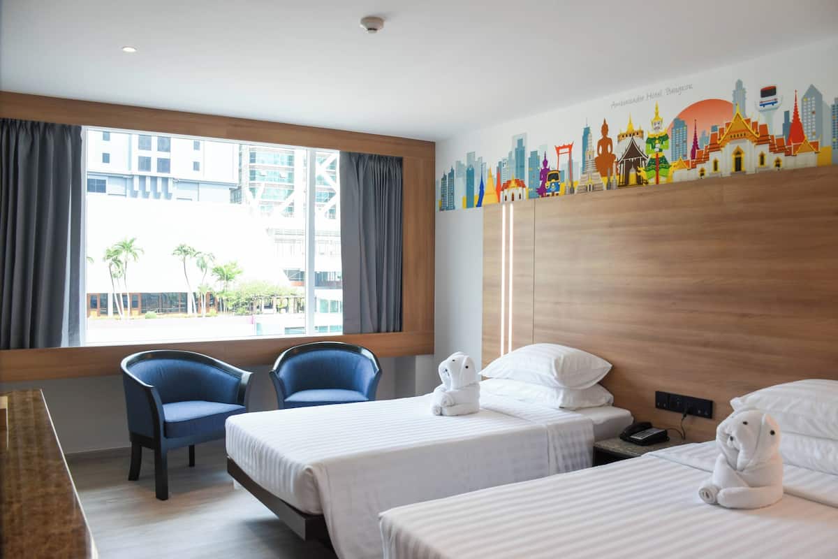 Quarto de hotel com duas camas, uma mesa, uma cadeira, um mural com paisagem urbana e vista para palmeiras e edifícios.
