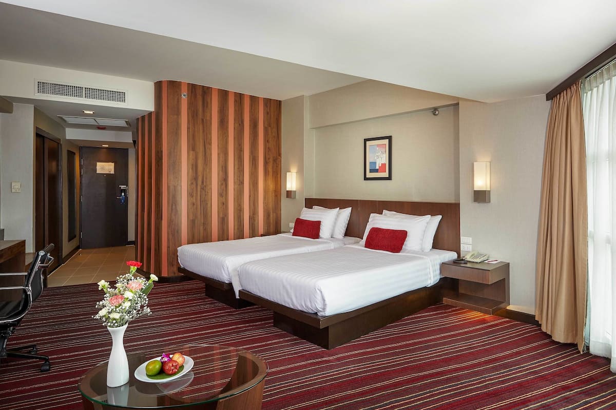 Quarto de hotel com duas camas, uma mesa de escritório com cadeira, uma mesinha com frutas e um vaso de flores.