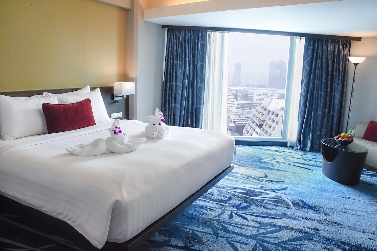 Quarto de hotel com uma cama grande, vista para a cidade e uma mesinha de cabeceira com uma fruteira.