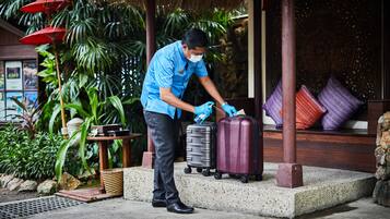 Funcionário com uniforme azul manuseando malas na entrada de um hotel, com plantas tropicais e estrutura de madeira.