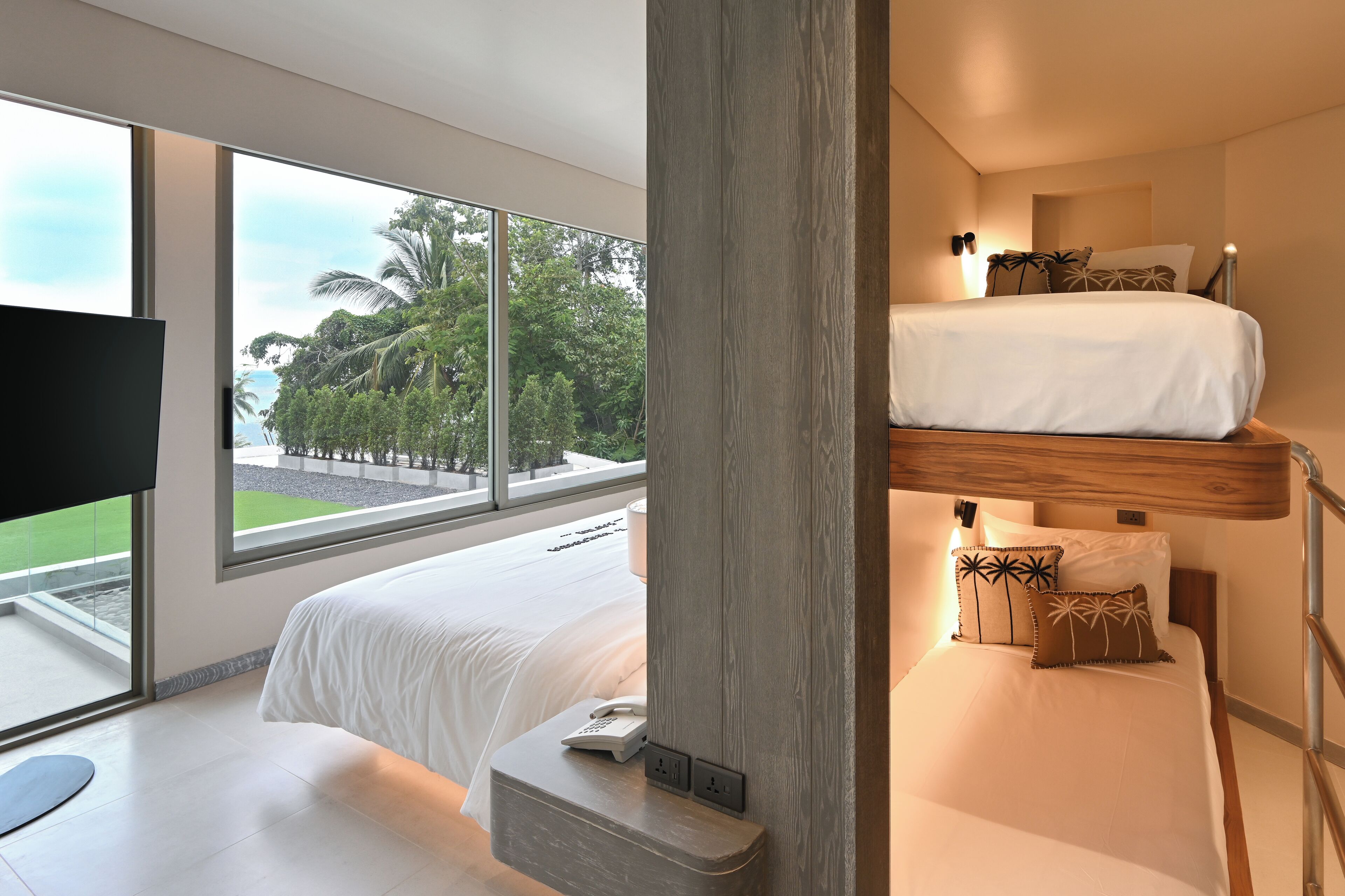 Quarto de hotel moderno com uma cama grande, televisão e vista para palmeiras através de amplas janelas.