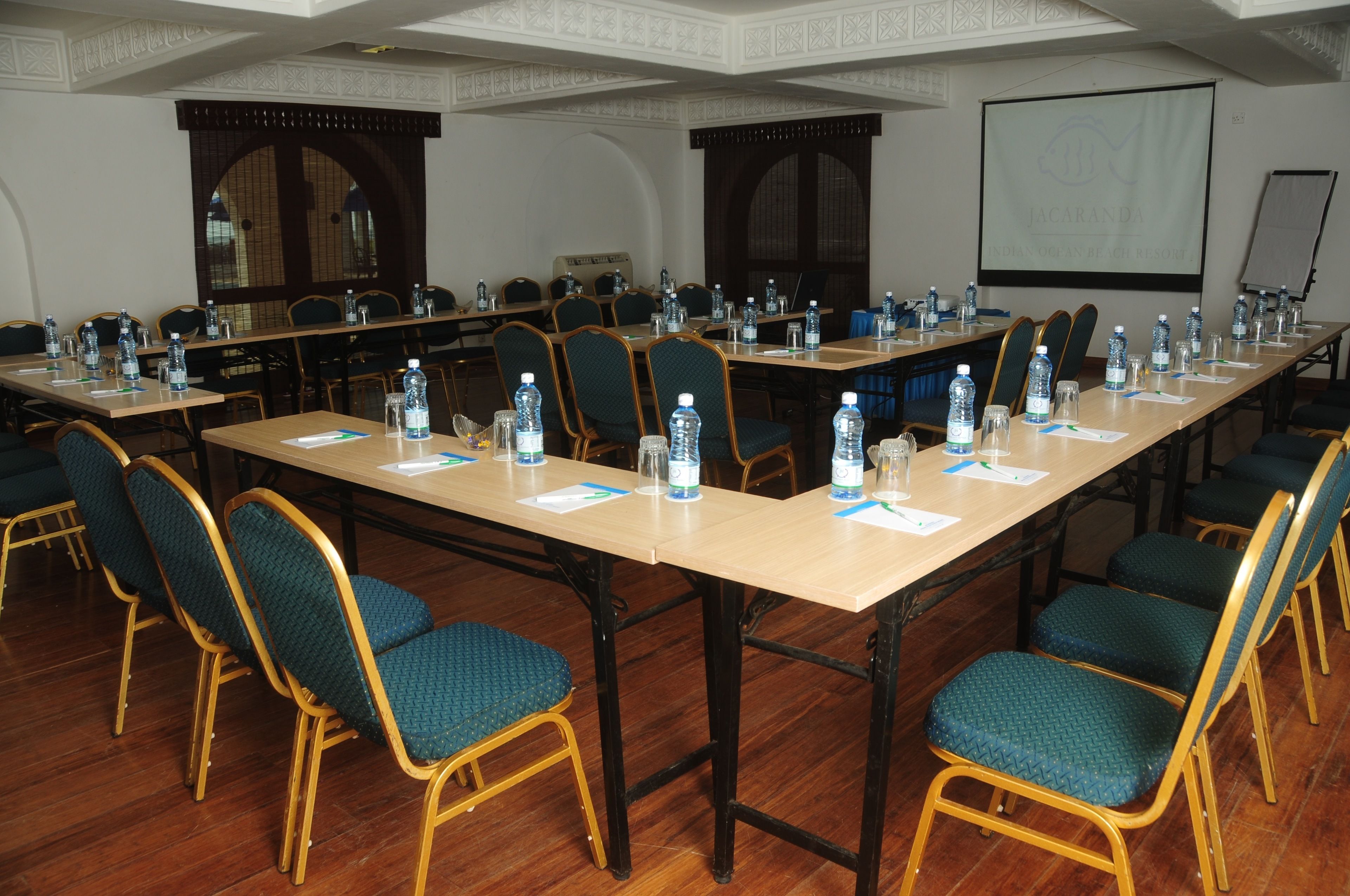 Uma sala de conferências com mesas grandes, cadeiras, garrafas de água e um projetor.