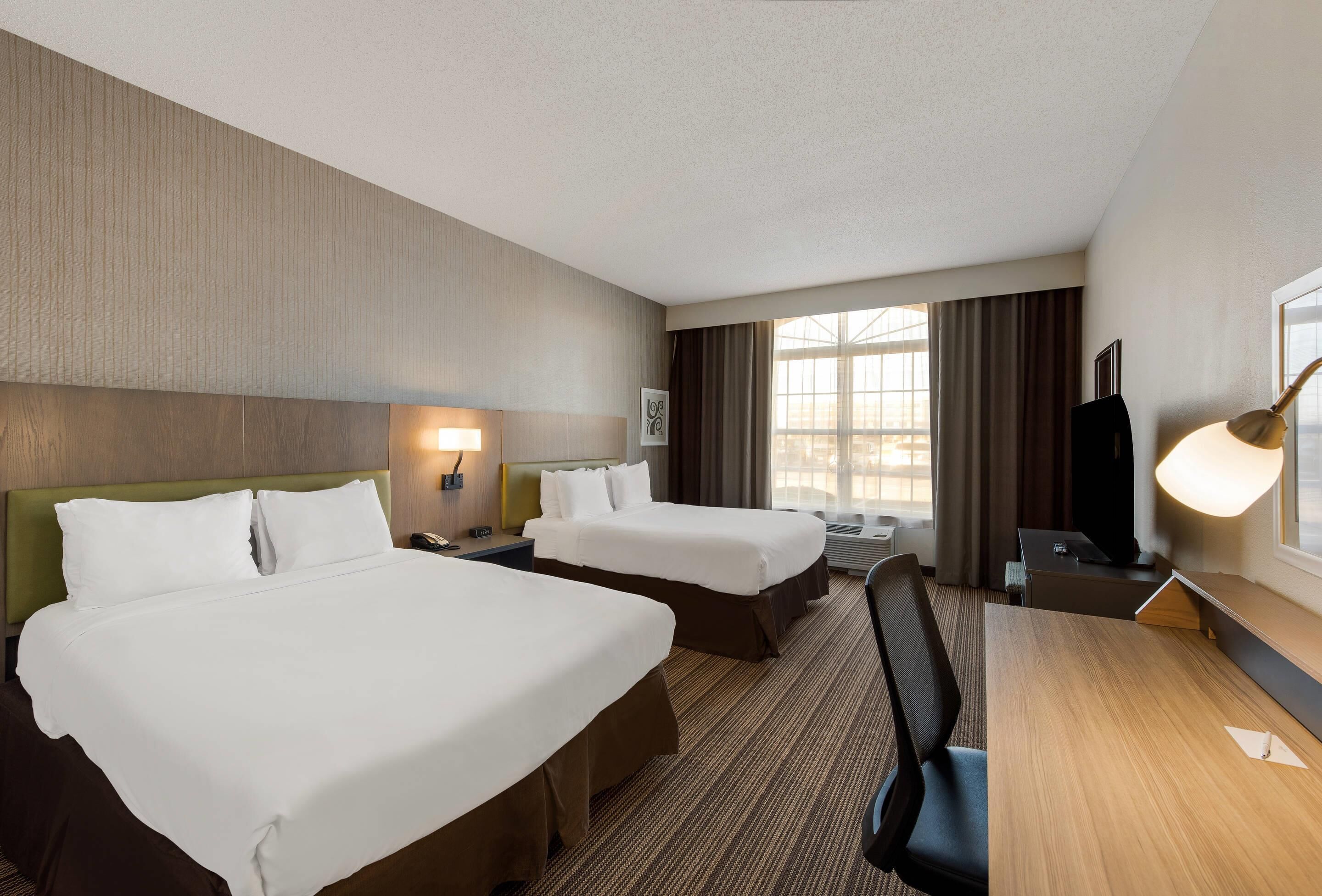 Quarto de hotel com duas camas, uma escrivaninha, uma cadeira, uma televisão e uma janela com cortinas.
