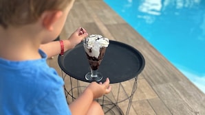 Criança sentada à mesa junto à piscina, com uma sobremesa.