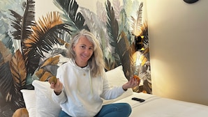 Uma pessoa sentada na cama com uma bandeja de itens de café da manhã, incluindo torradas, frutas e um copo de suco de laranja.