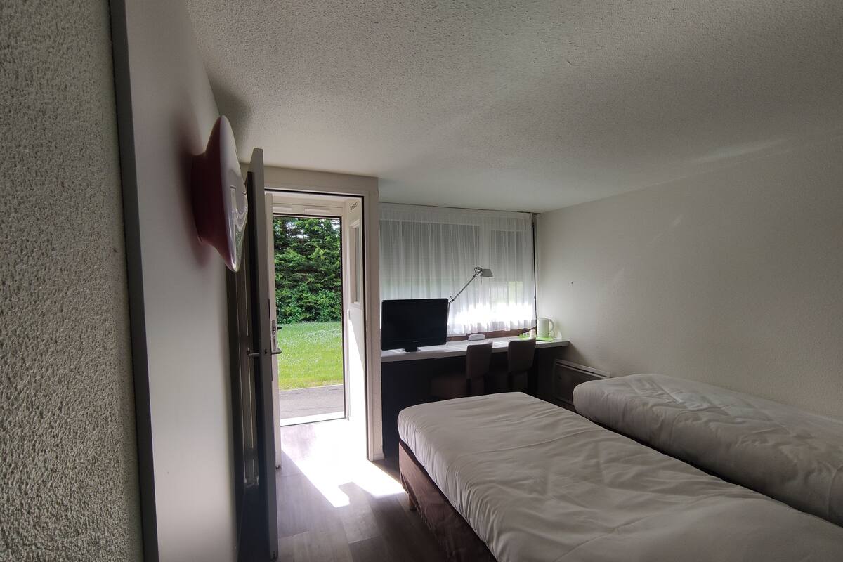 Quarto de hotel com uma cama, uma escrivaninha com computador, uma cadeira e uma janela com cortinas.