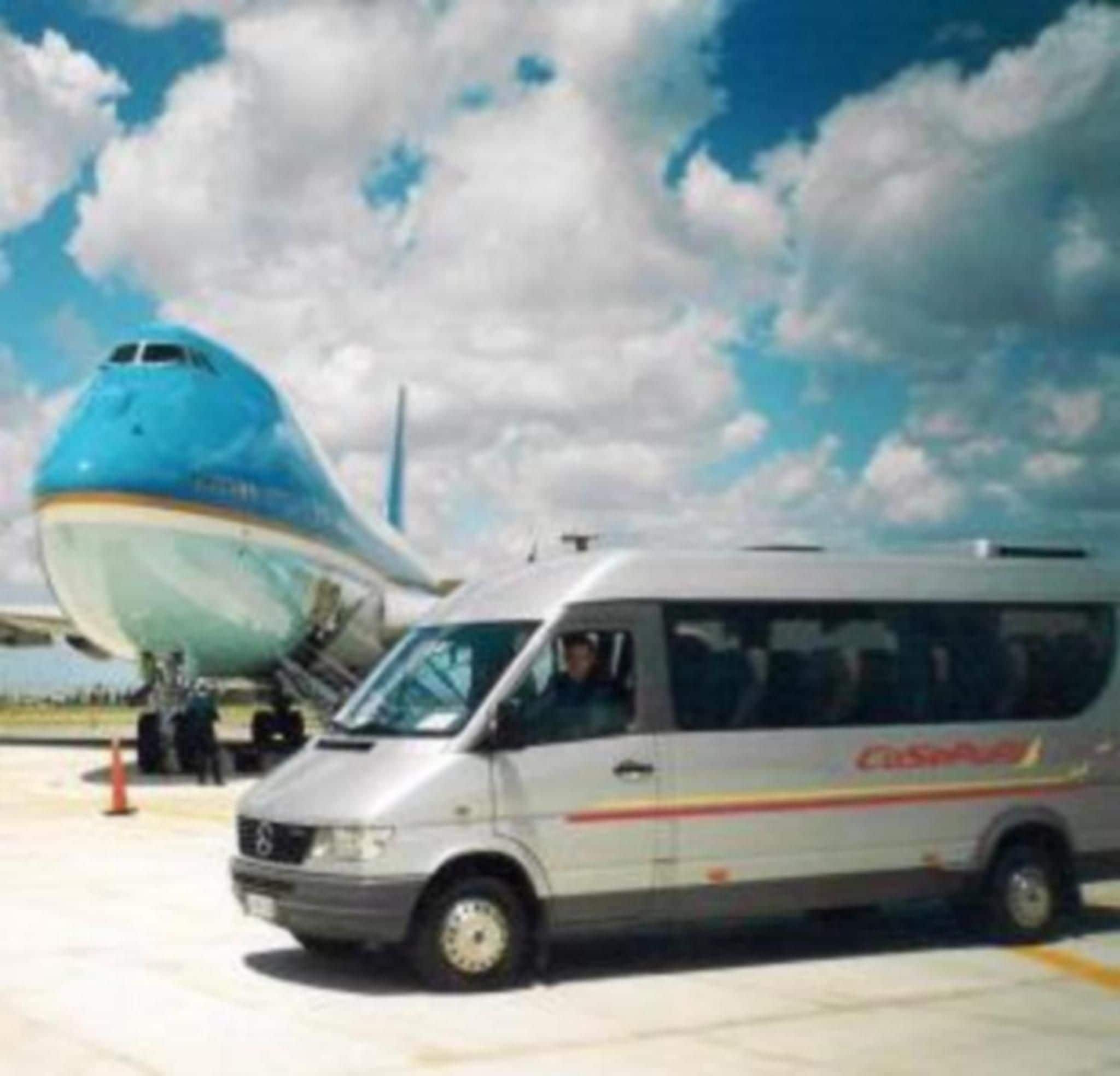 Um ônibus estacionado no pavimento de um aeroporto, com a ponta de um avião visível ao fundo.