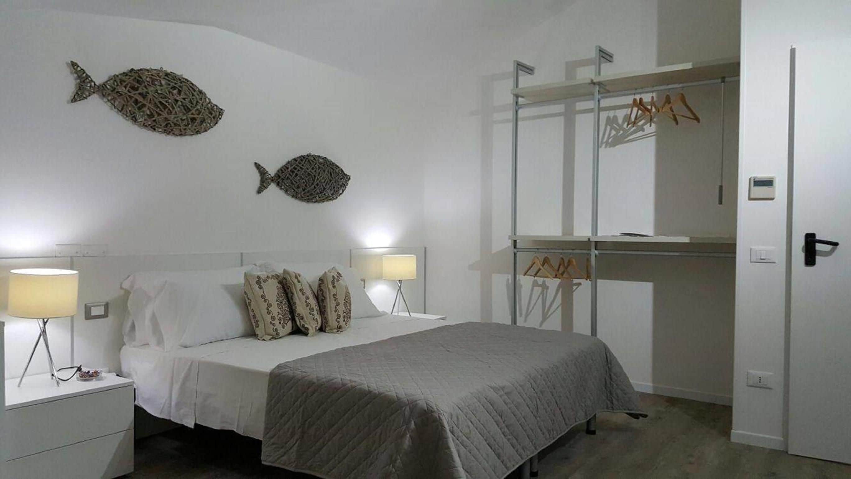 Quarto com cama, duas luminárias fixadas na parede, mesa de cabeceira, guarda-roupa com espelho e duas decorações de peixes na parede.