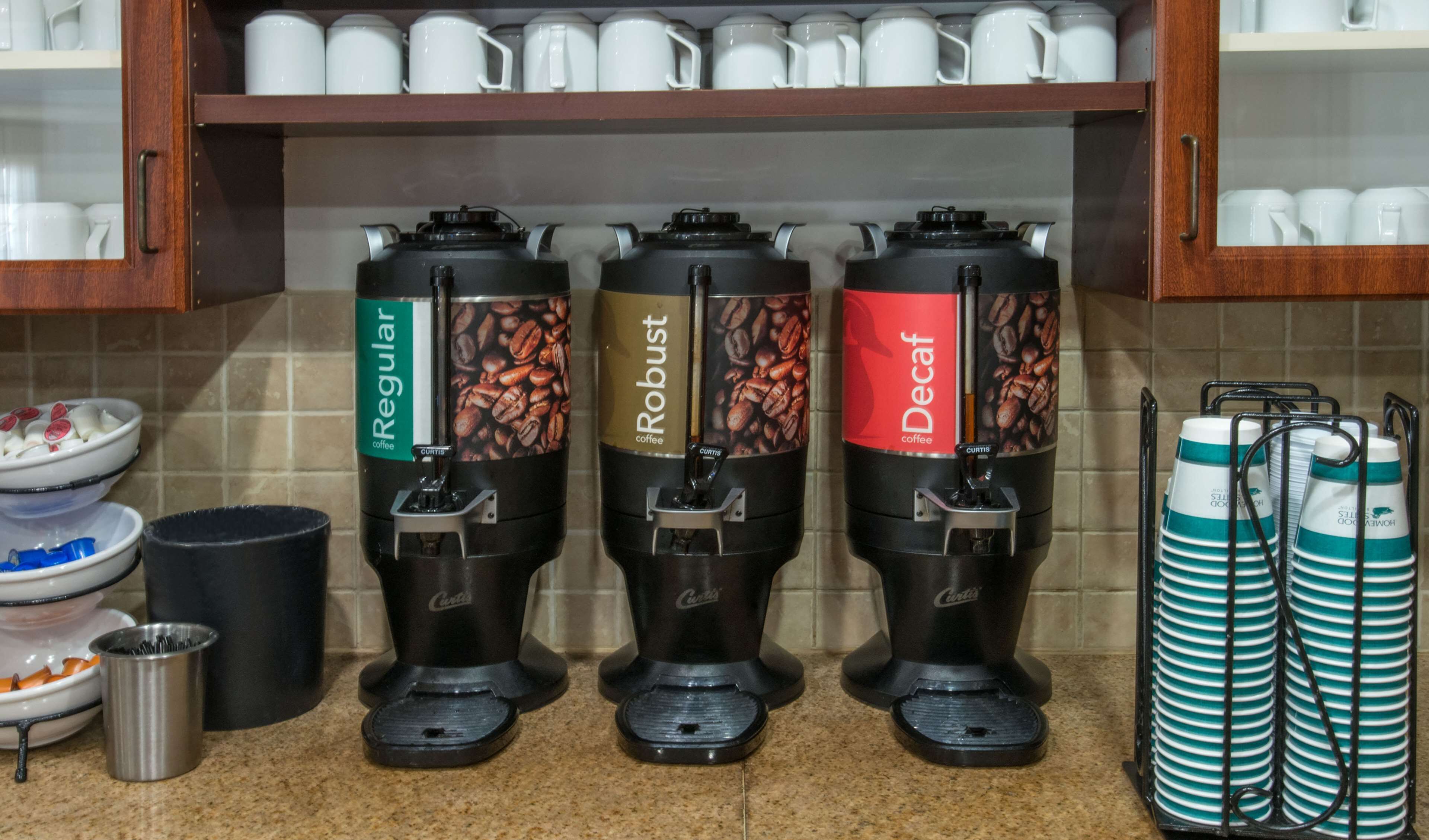 Três dispensadores de café identificados como Tradicional, Intenso e Descafeinado em uma cozinha, com uma cafeteira e xícaras sobre o balcão.