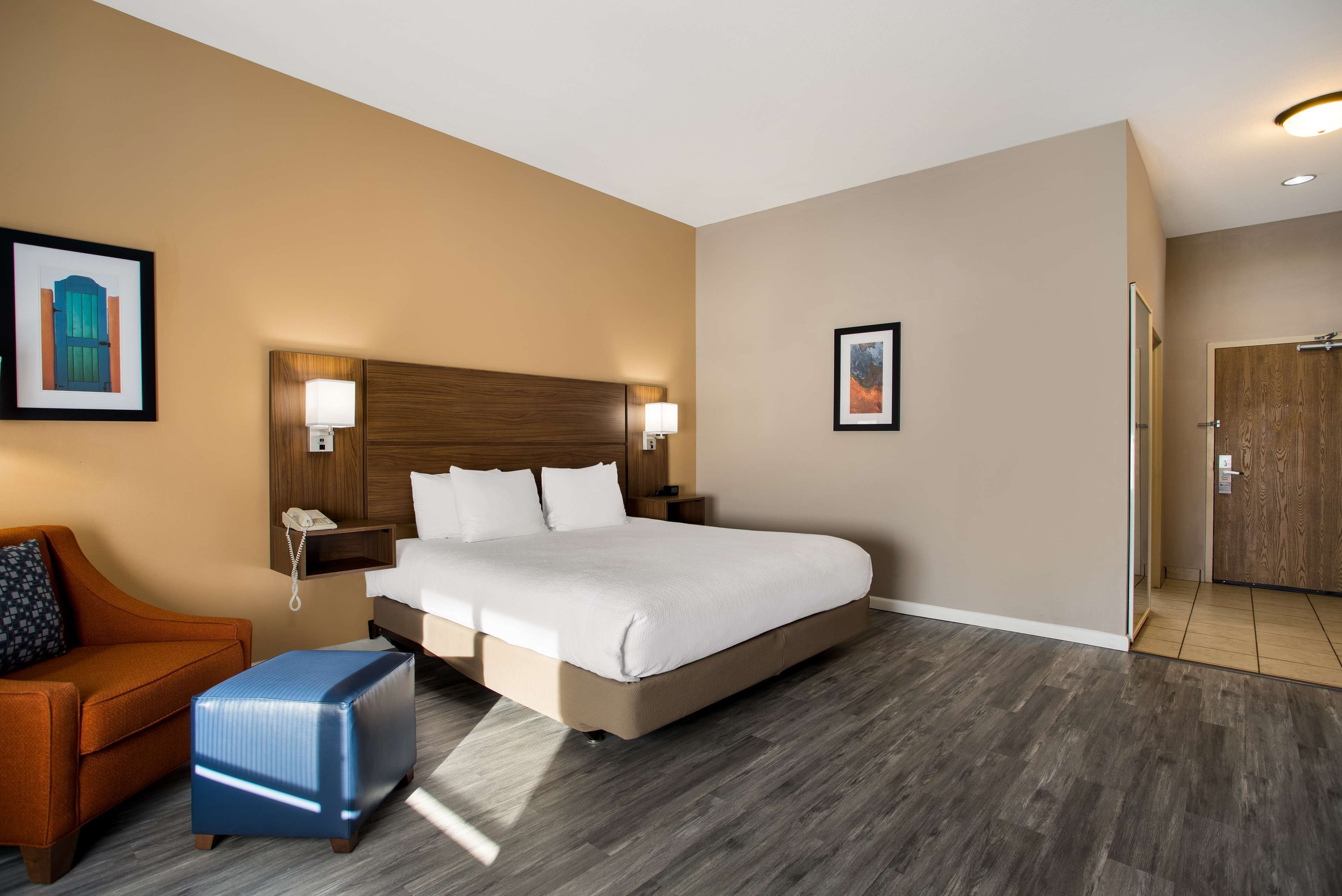 Quarto de hotel com uma cama grande, uma mesa de cabeceira com abajur, telefone, um banquinho azul e um quadro na parede.