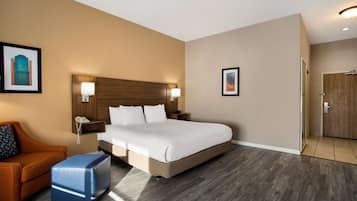 Quarto de hotel com uma cama grande, uma mesa de cabeceira com abajur, telefone, um banquinho azul e um quadro na parede.