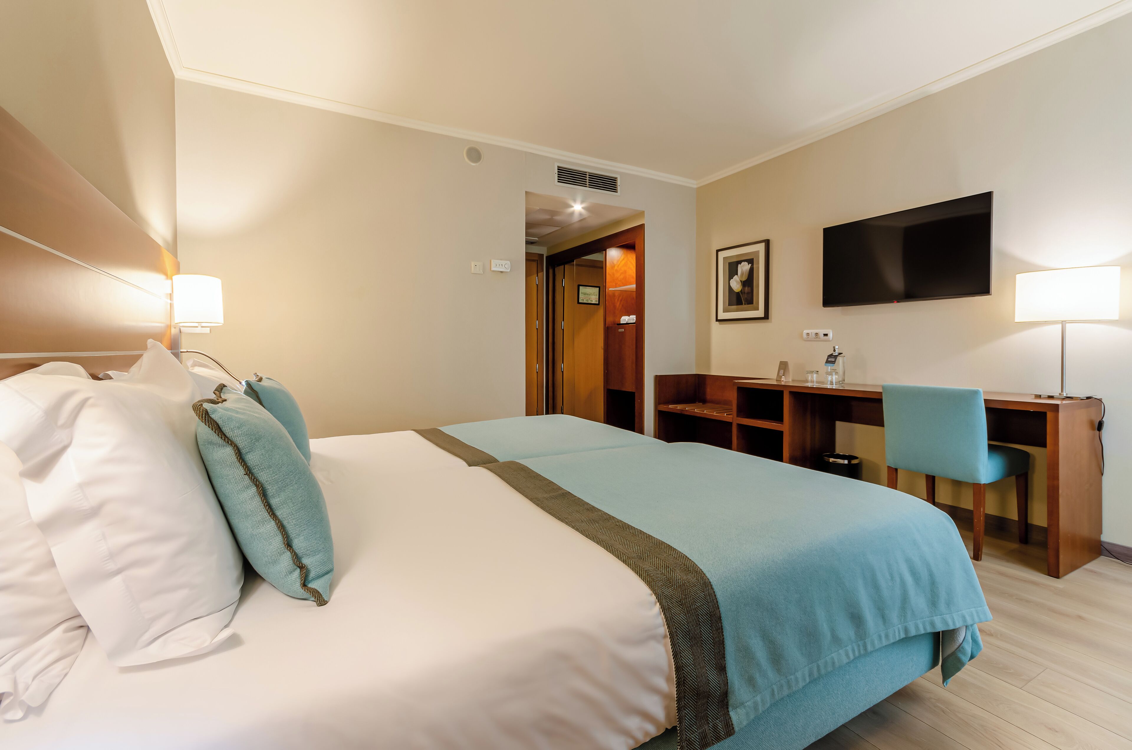 Quarto de hotel com cama, escrivaninha, cadeira e televisão.