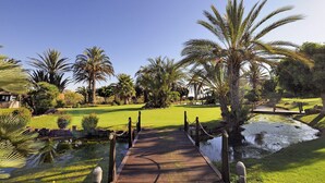 Uma ponte de madeira que dá acesso a um campo de golfe, com um lago e palmeiras.