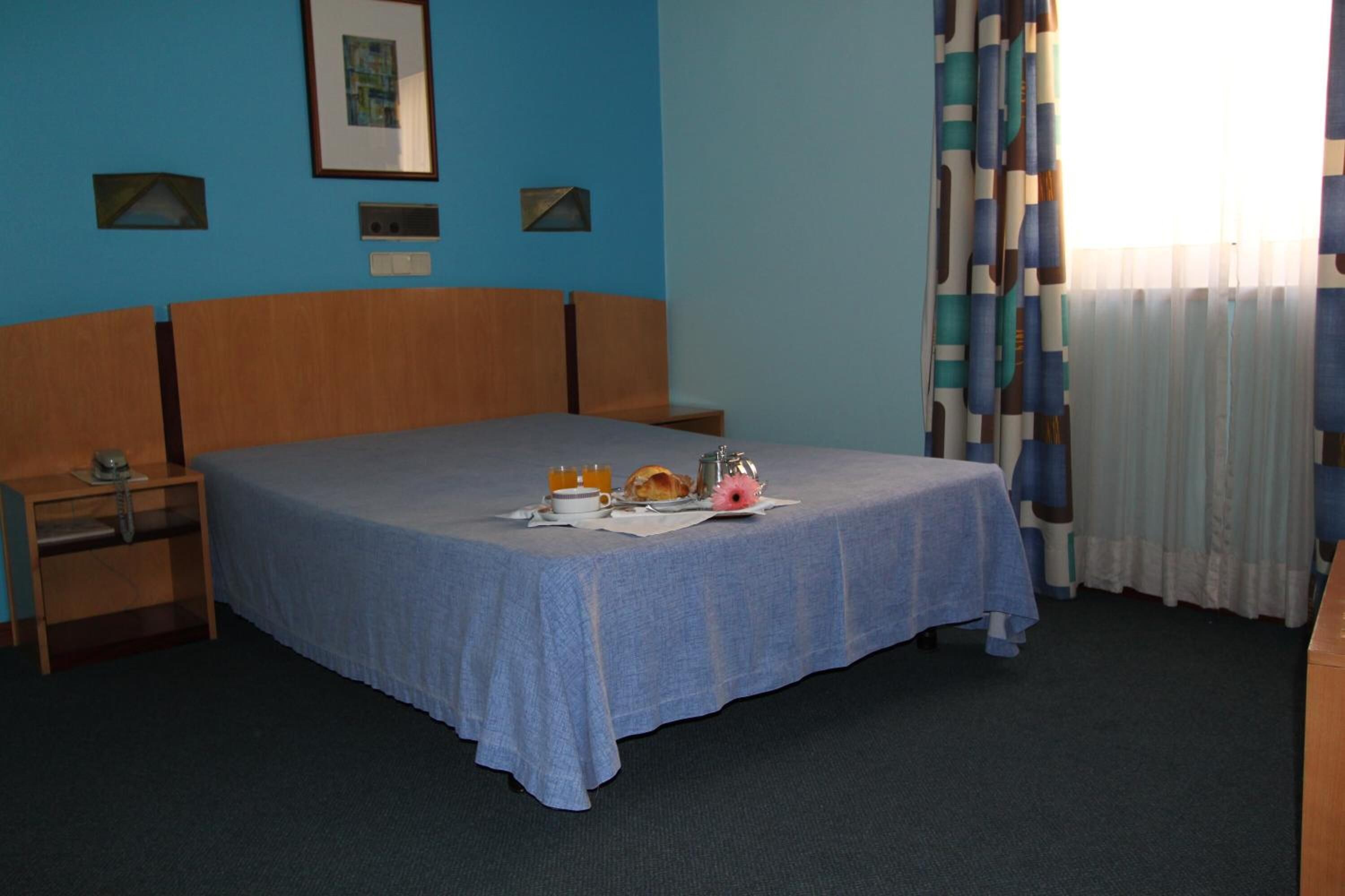 Quarto de hotel com cama, mesa de cabeceira e janela com cortinas.