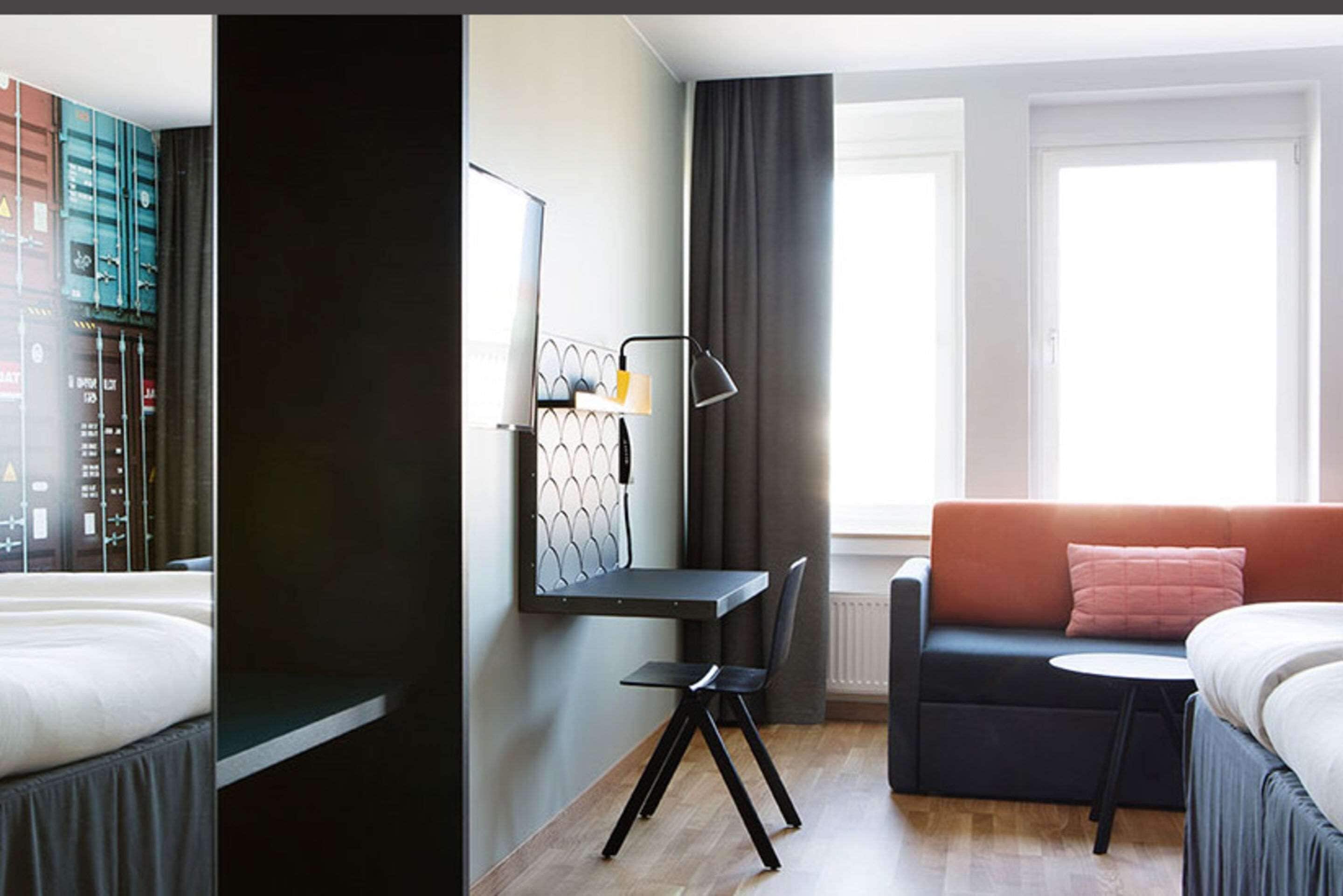 Quarto de hotel com uma cama, uma escrivaninha com cadeira, um sofá e uma janela com cortinas.