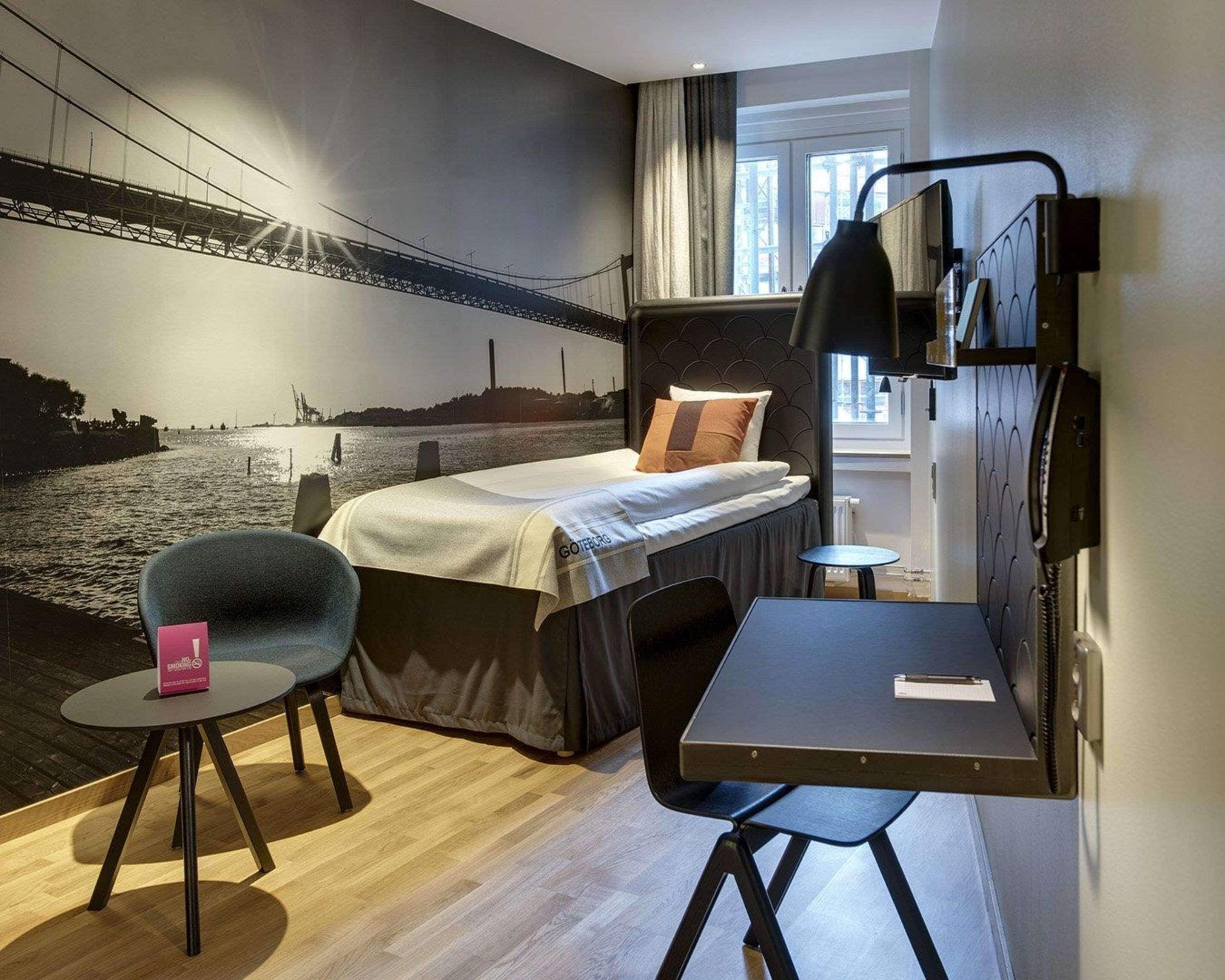 Quarto de hotel pequeno, com uma cama, uma escrivaninha, uma cadeira e um mural na parede de uma ponte.