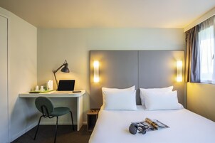 Quarto de hotel com cama, escrivaninha com um laptop, cadeira e mesa de cabeceira.