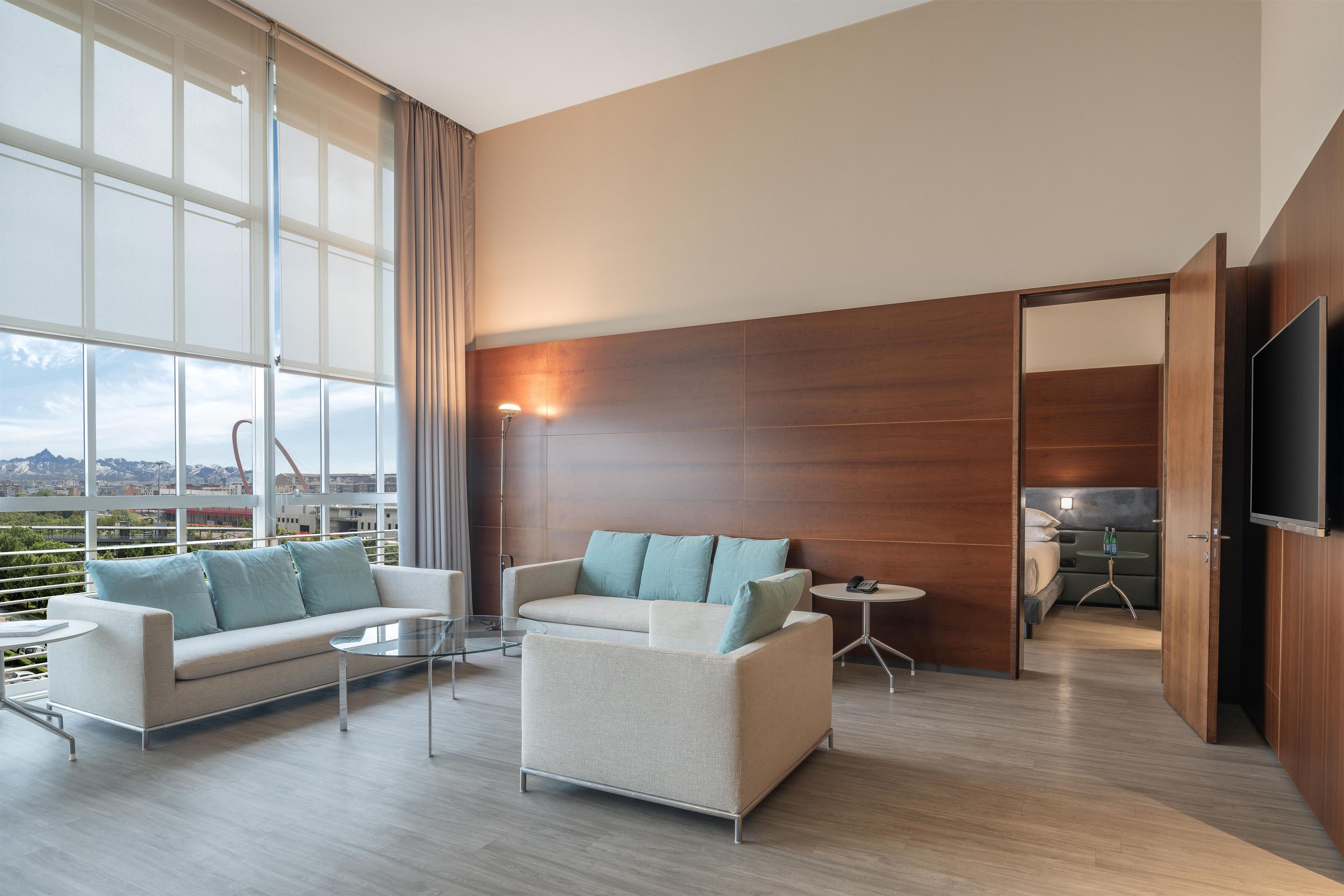 Quarto de hotel moderno com uma janela grande, um sofá, uma mesa de centro e uma TV fixada na parede.